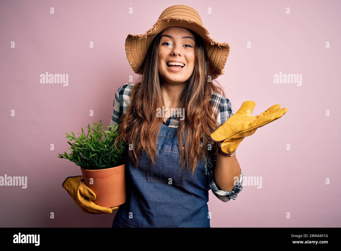 Young beautiful brunette farmer woman wearing apron and hat holding pot ...