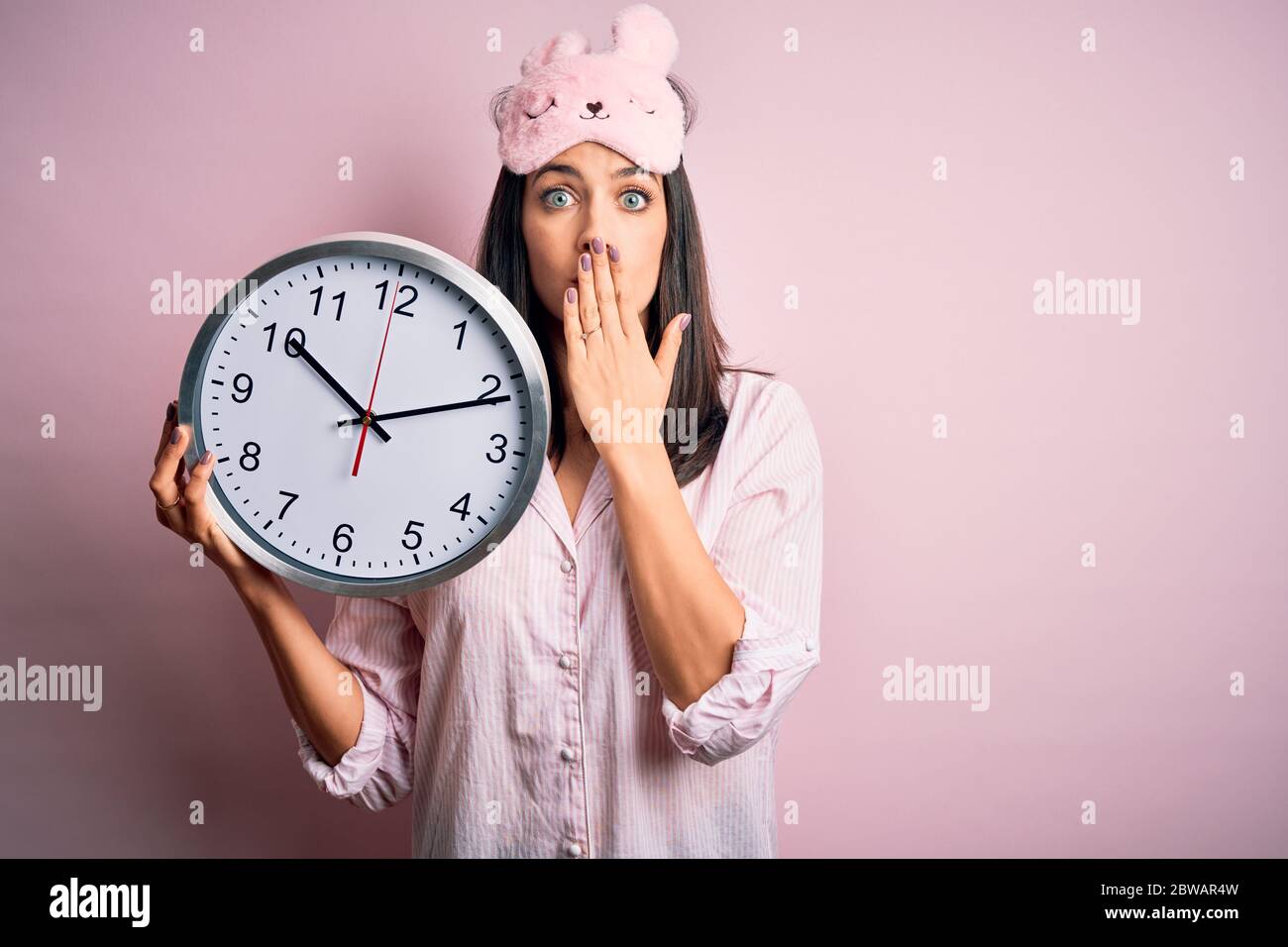Young brunette woman with blue eyes wearing pajama holding big clock at ...