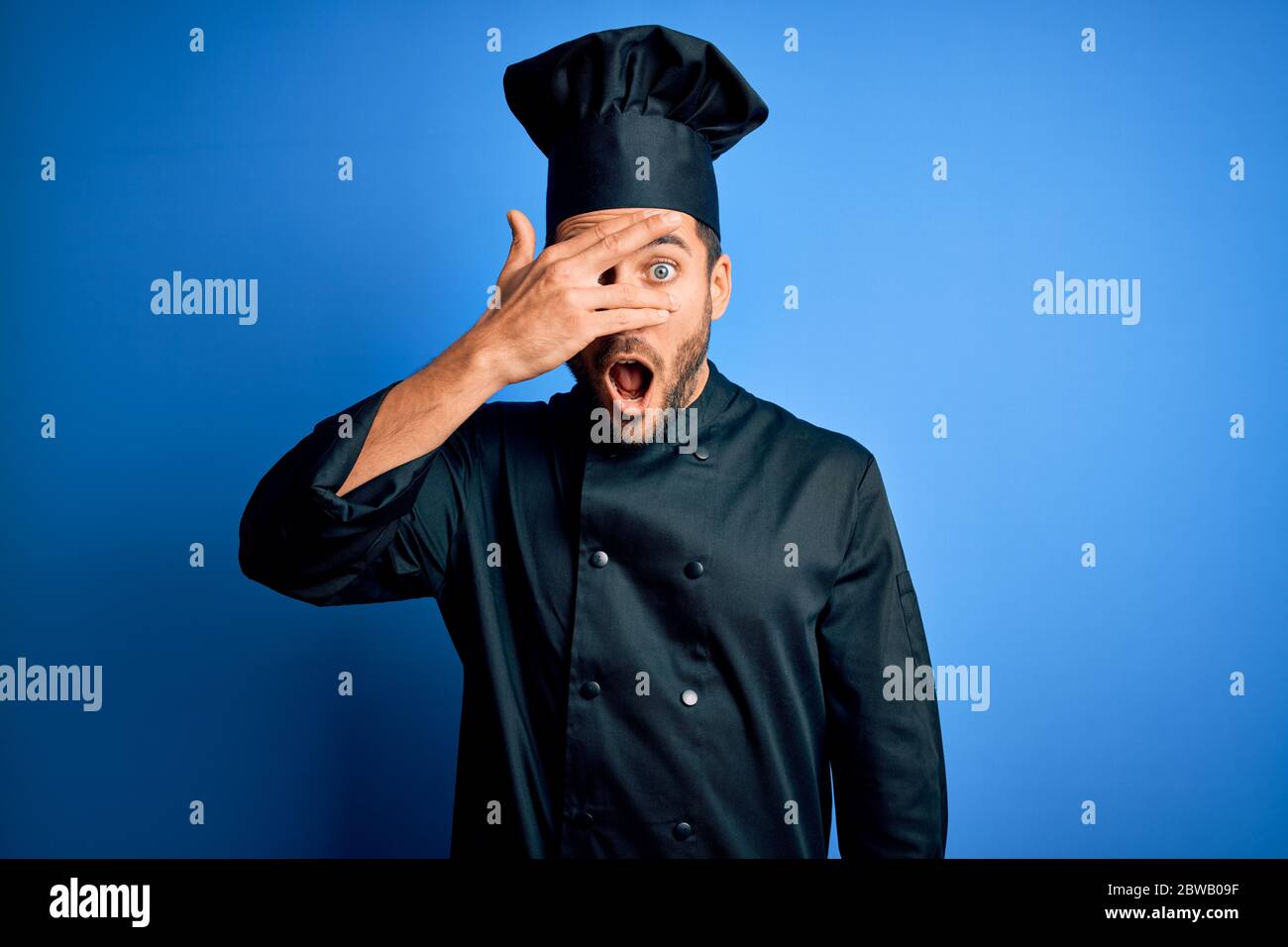 Young handsome chef man with beard wearing cooker uniform and hat over ...