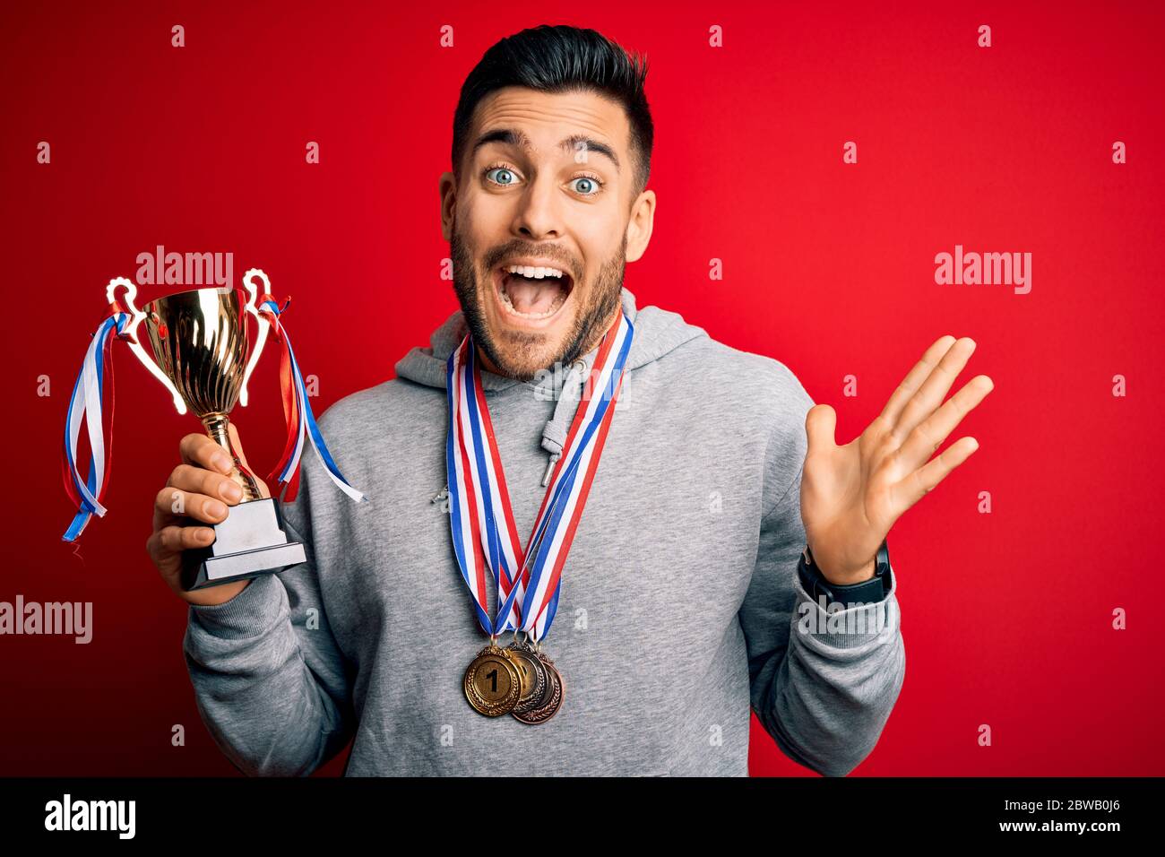 Young handsome succesful man holding trophy wearing medals over red ...