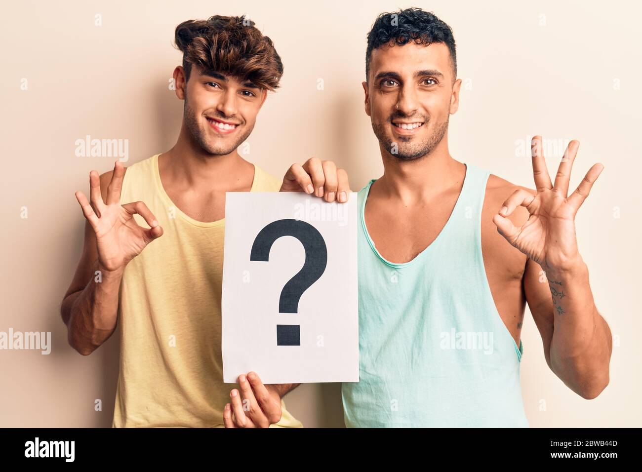 Young gay couple holding question mark doing ok sign with fingers ...