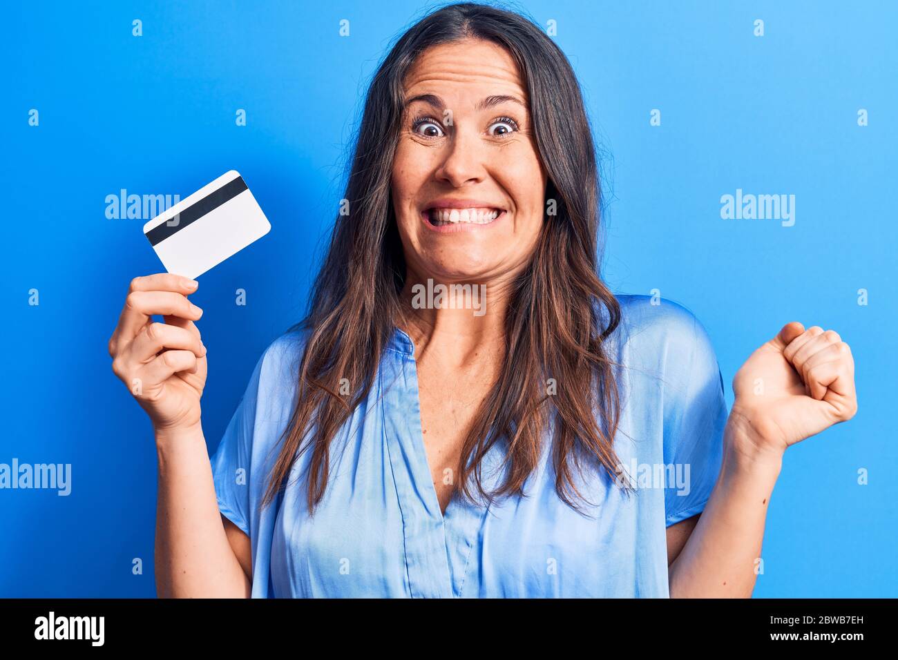 Young beautiful brunette woman holding credit card over isolated blue ...