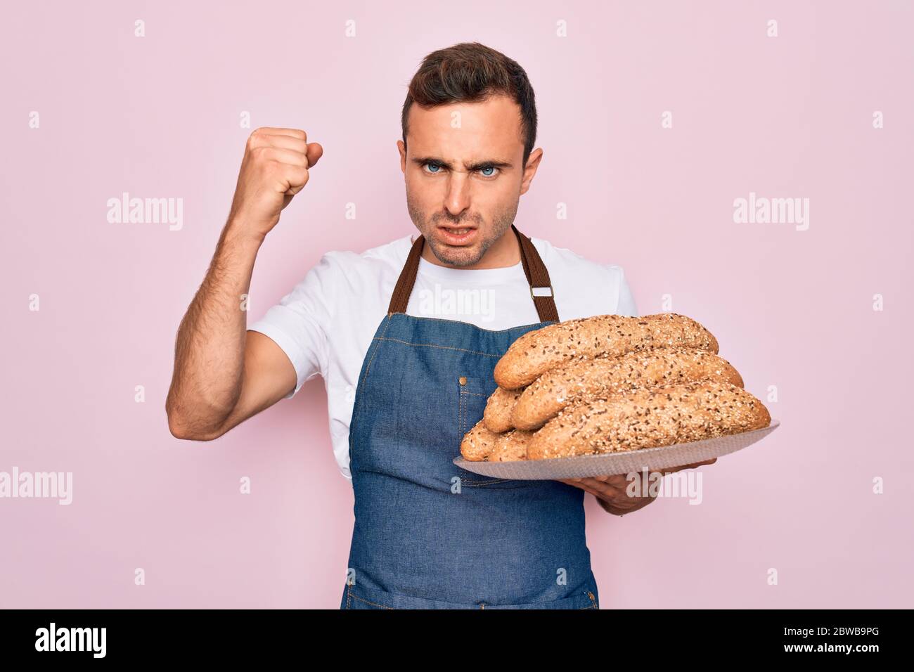 Young handsome baker man with blue eyes wearing apron holding tray with ...