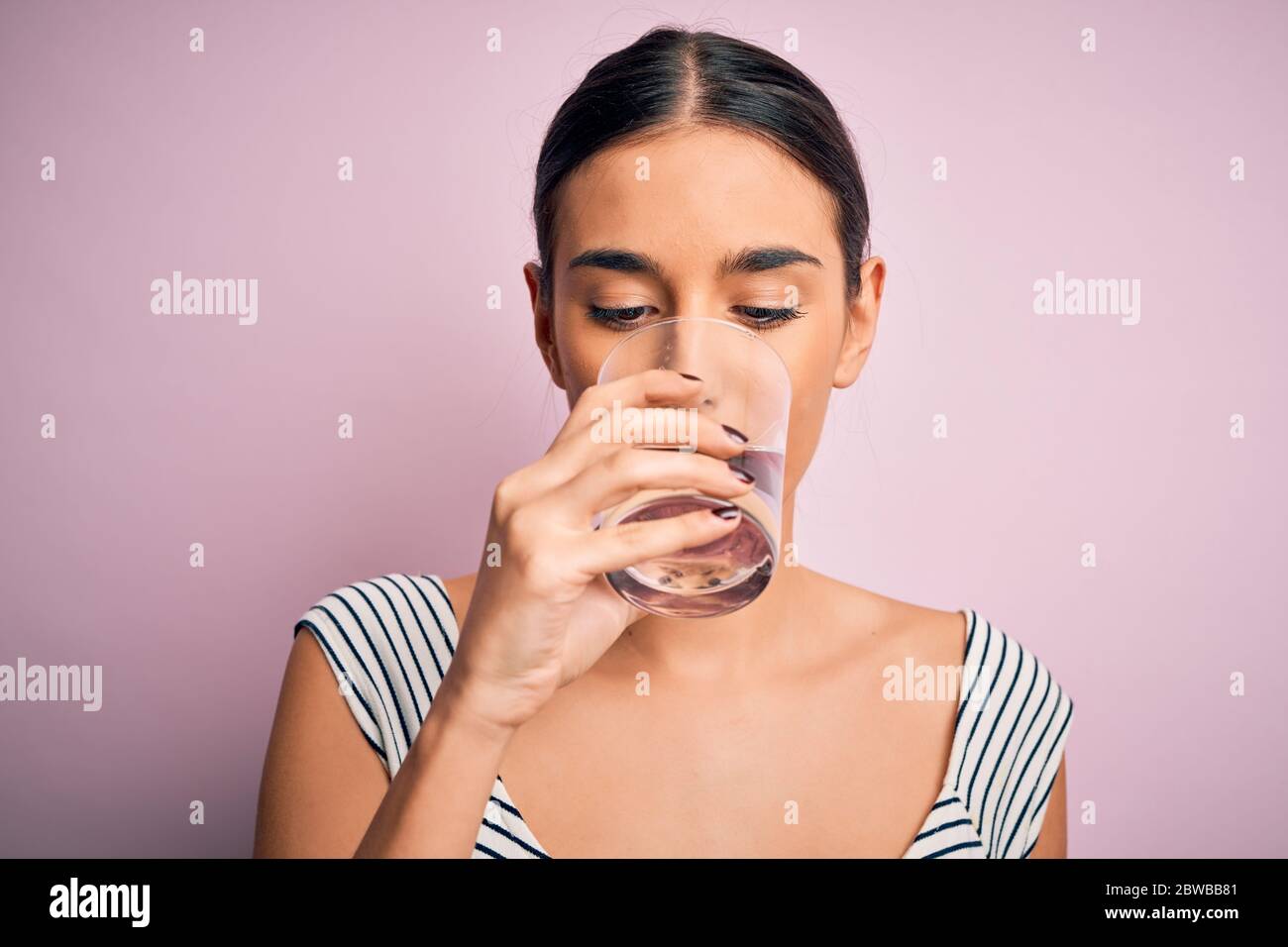 Young beautiful brunette woman drinking glass of healthy water to refreshment standing over ...