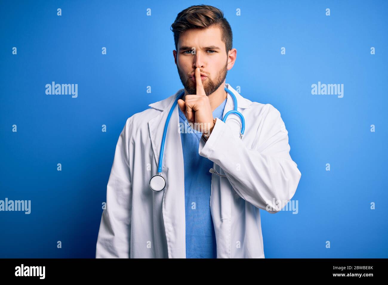 Young blond doctor man with beard and blue eyes wearing white coat and ...
