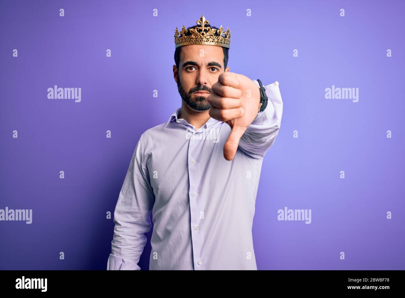 Young handsome man with beard wearing golden crown of king over purple ...