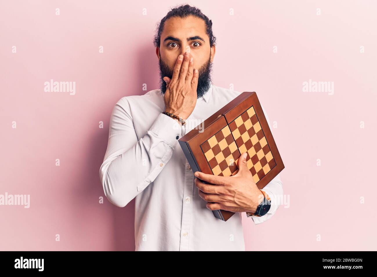 Young arab man holding chess covering mouth with hand, shocked and ...