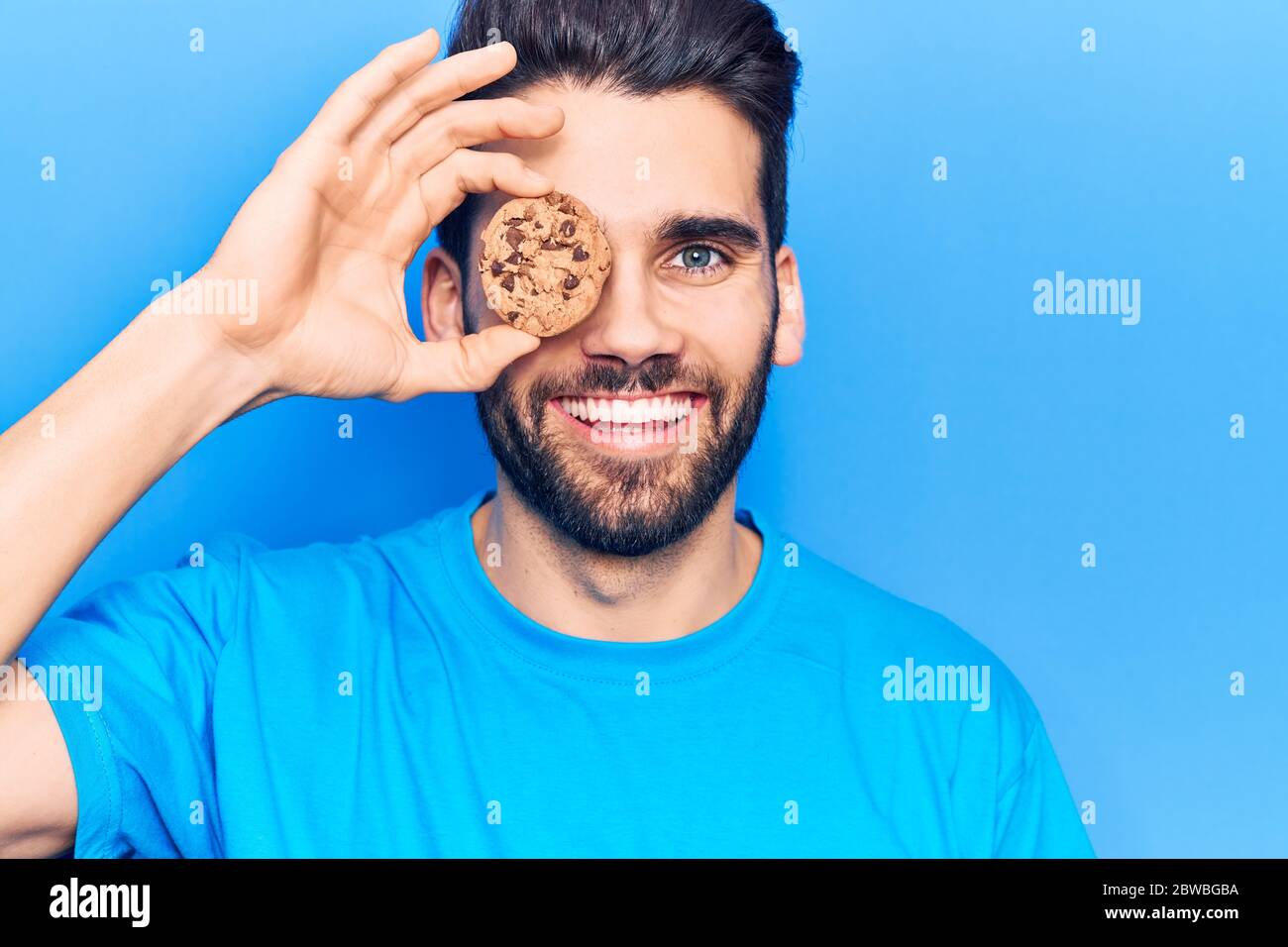 Young handsome man with beard holding chocolate cookie looking positive ...