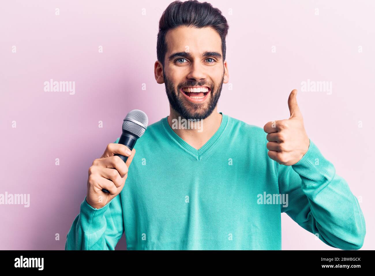 Young handsome man with beard singing song using microphone smiling happy and positive, thumb up ...