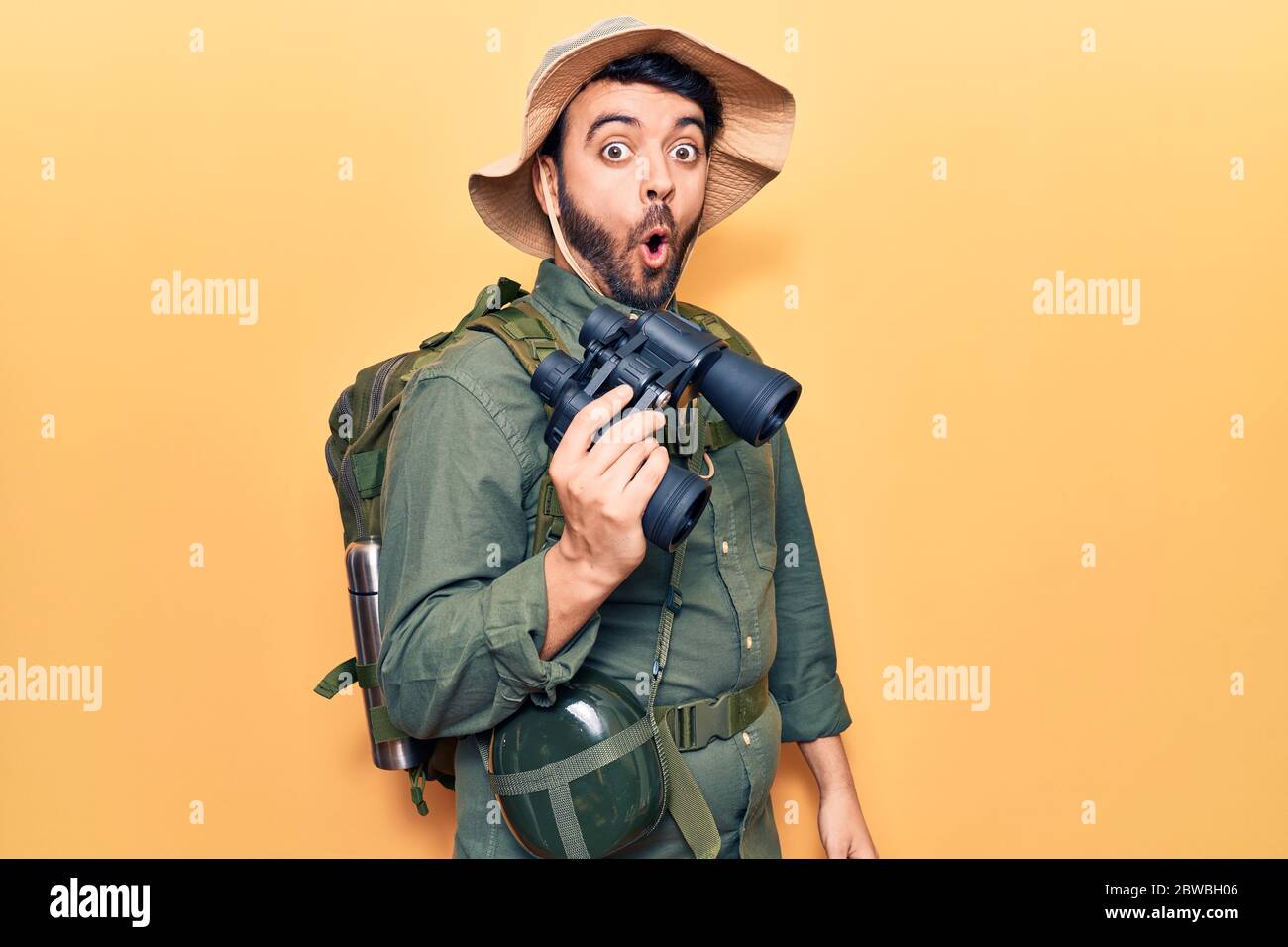 Young hispanic man wearing explorer clothes holding binoculars scared ...