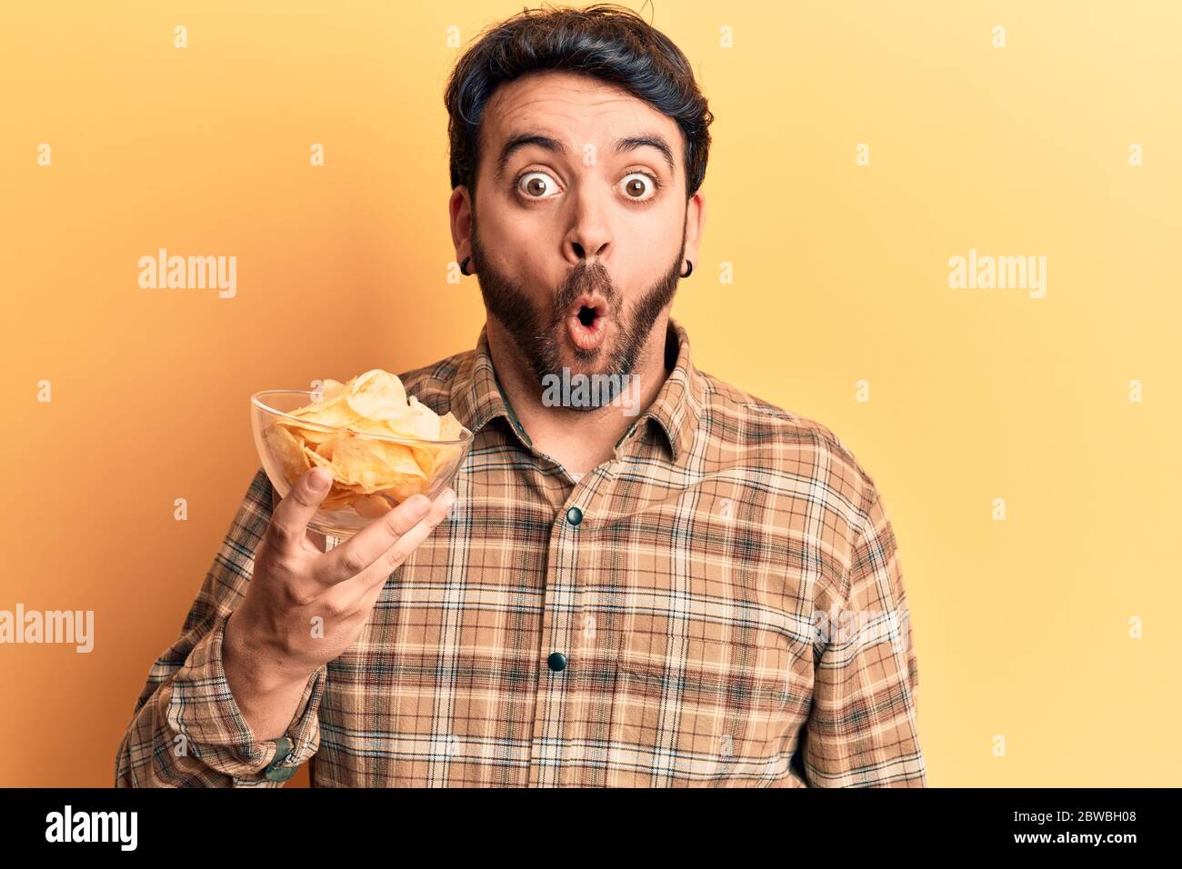 Young hispanic man holding potato chip scared and amazed with open ...