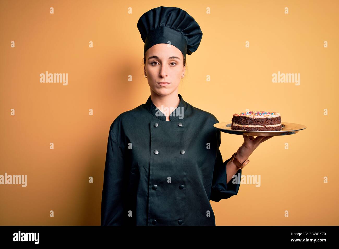 Young beautiful baker woman wearing cooker uniform and hat holding tray ...