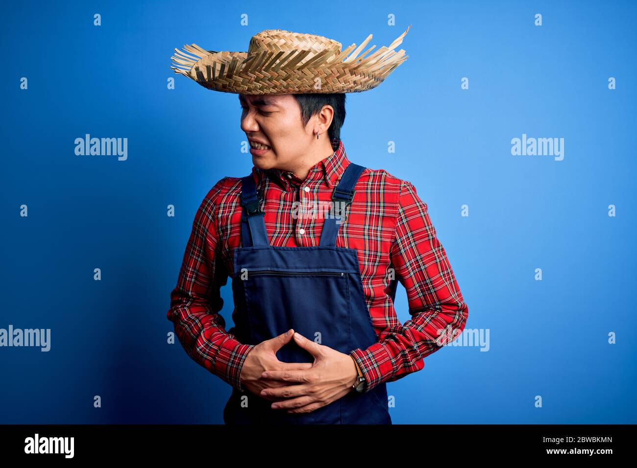 Young handsome chinese farmer man wearing apron and straw hat over blue ...