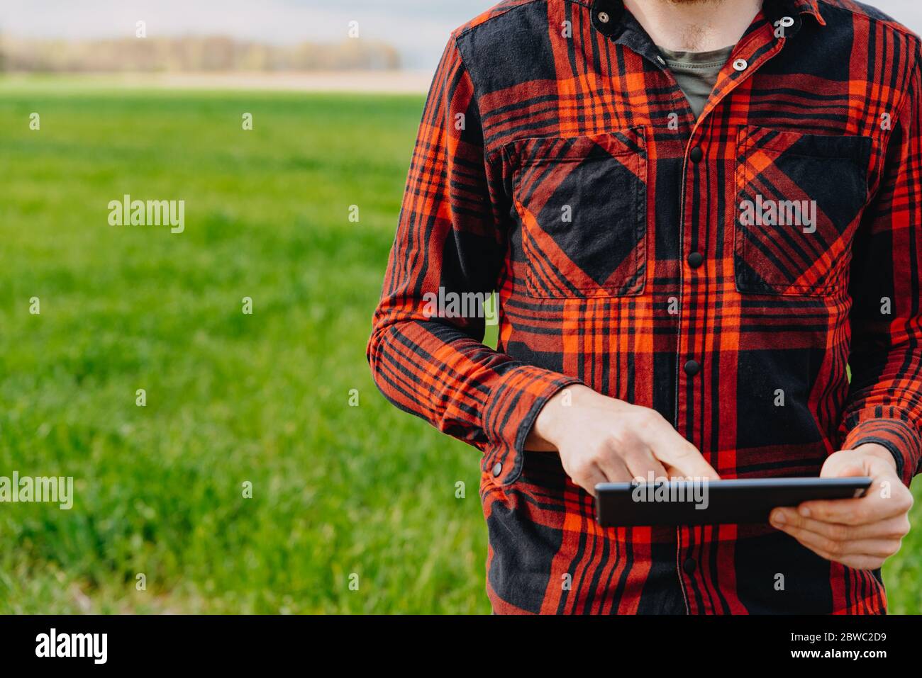 Farmer in red checked shirt using tablet on green wheat field. Applying ...