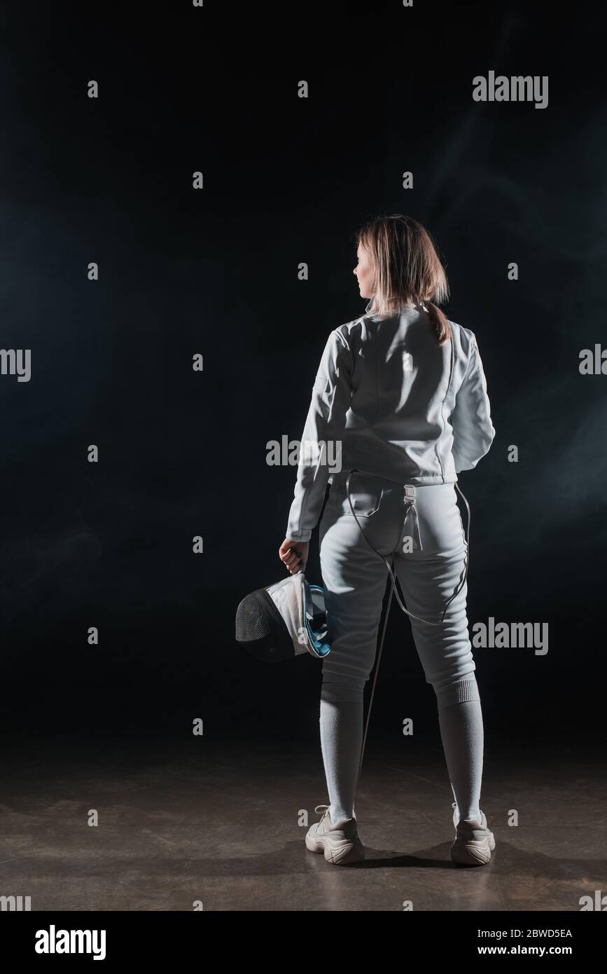 Back view of swordswoman holding fencing mask and rapier on black ...