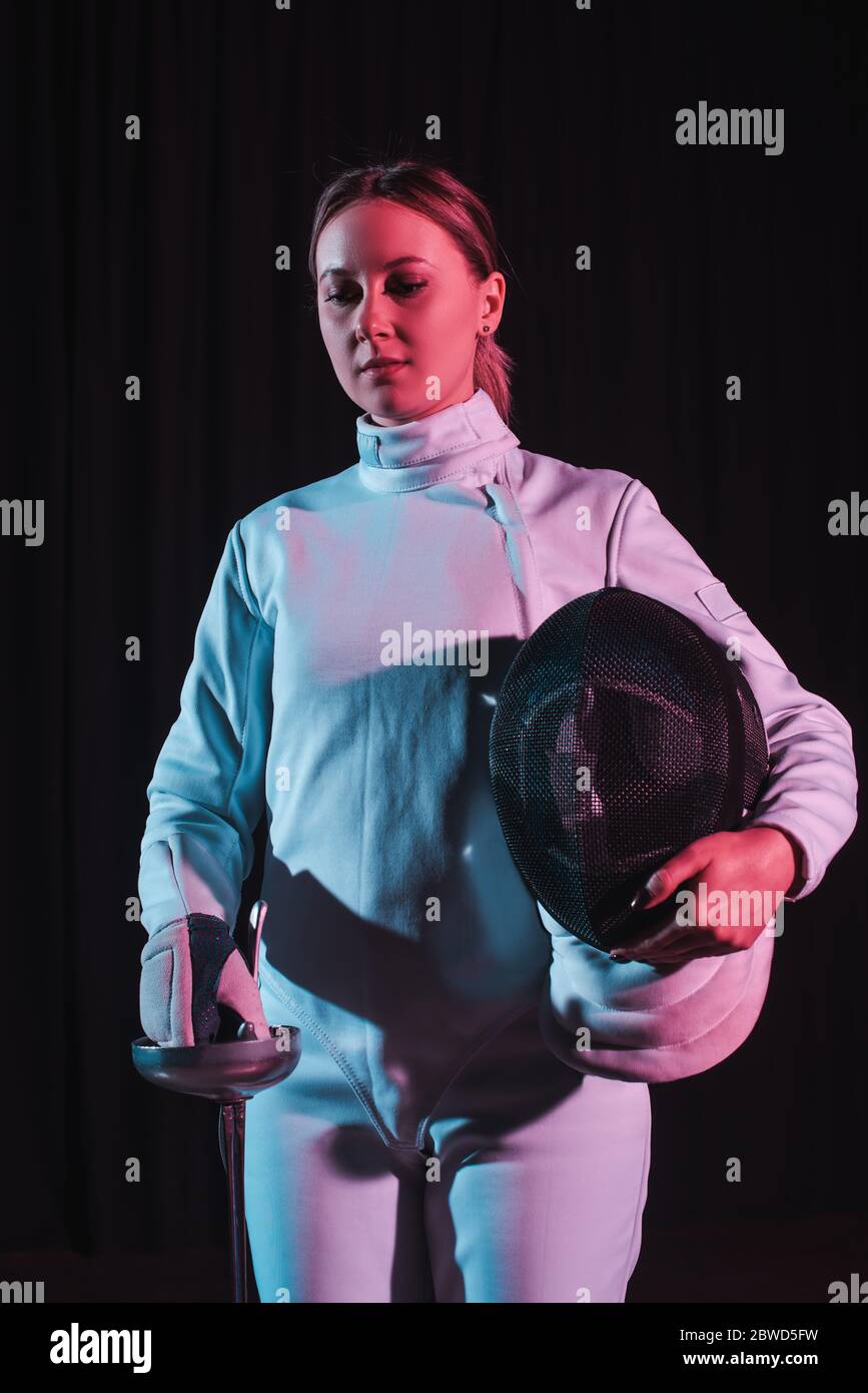 Attractive fencer holding fencing mask and rapier isolated on black ...