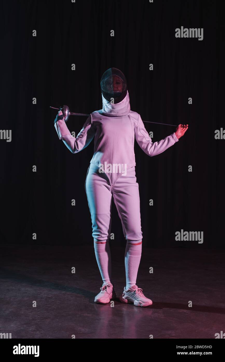 Fencer in fencing mask and suit holding rapier near shoulders on black ...
