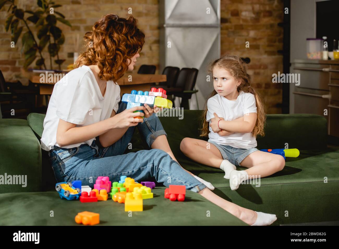 curly mother holding toy gun and looking at offended daughter with ...