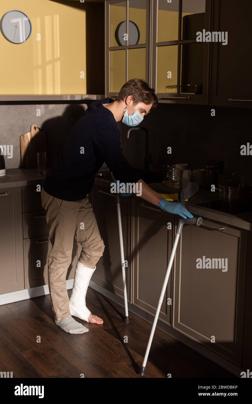 Side view of man with broken leg cleaning kitchen worktop near crutches ...