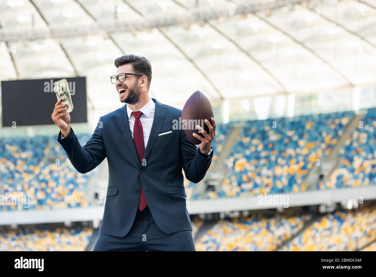 happy young businessman in suit and glasses with rugby ball and money ...