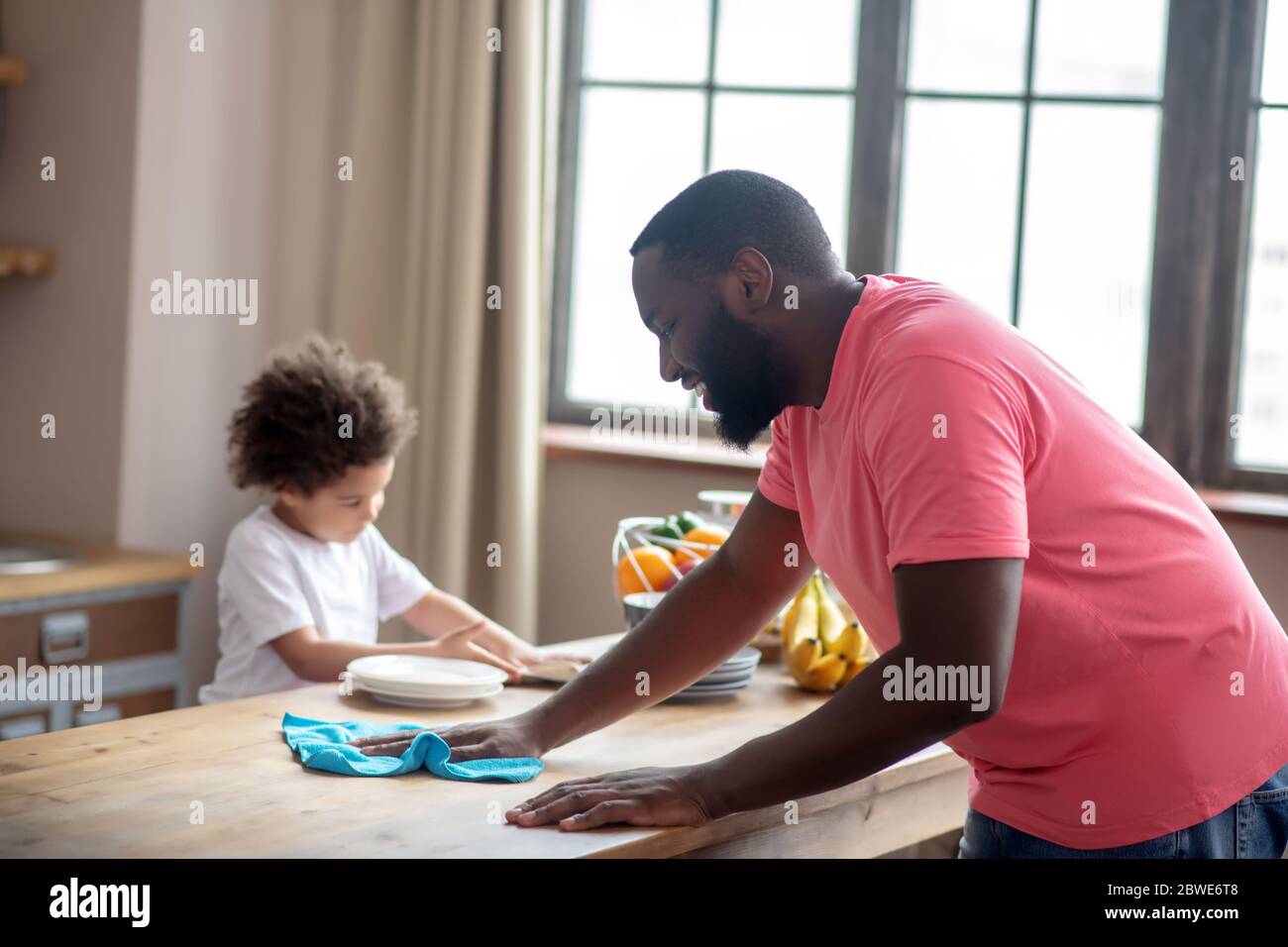 Tall dark-skinned man in a pink tshirt wiping the table while his kid ...