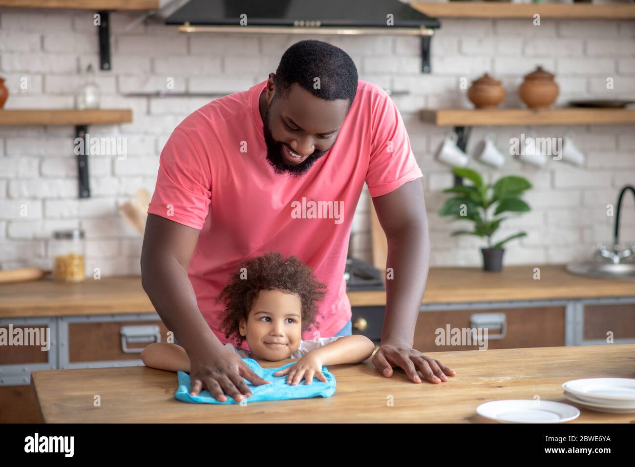 Tall dark-skinned man in a pink tshirt standing and wiping the table ...
