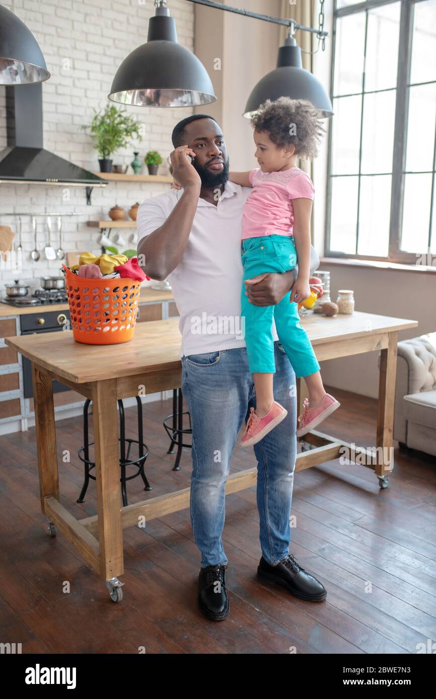 Dark-skinned tall father standing and holding his kid while talking on ...