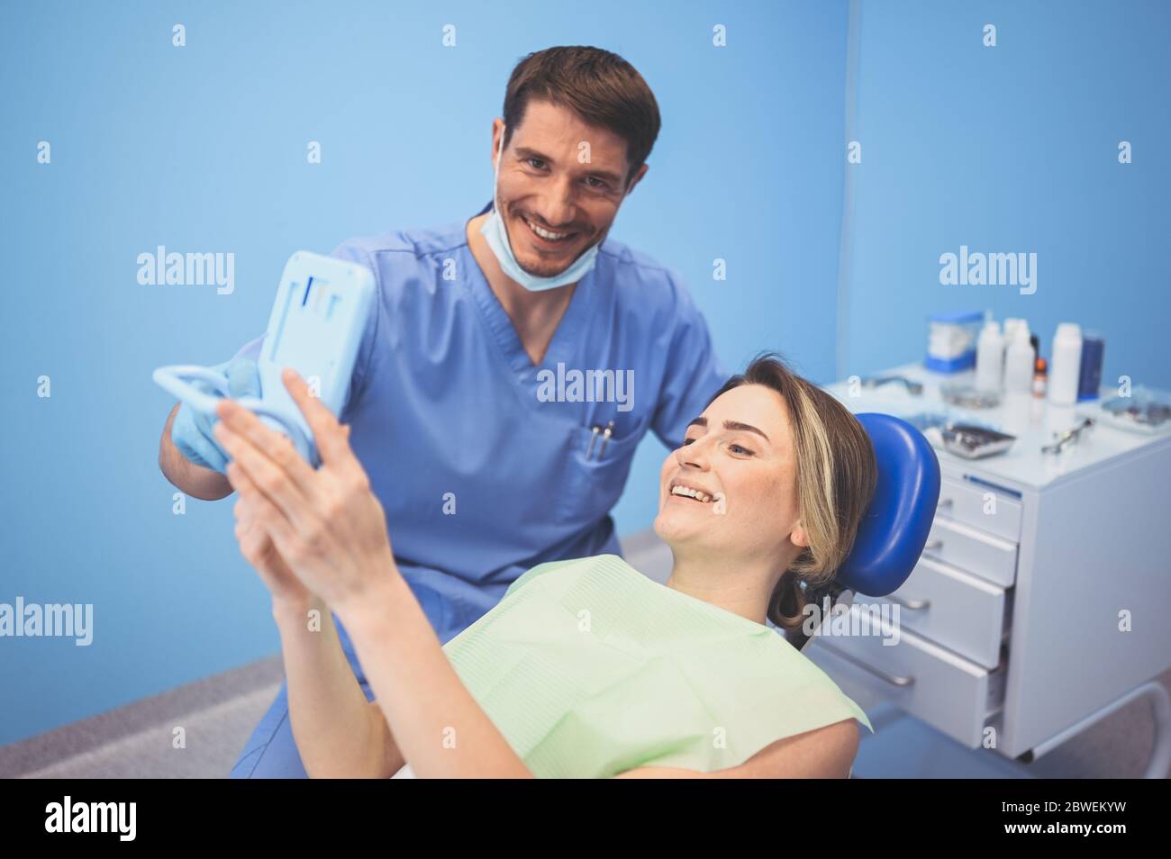 Dentist shows the patient the results of treatment with a mirror, examinating teeth with dental ...