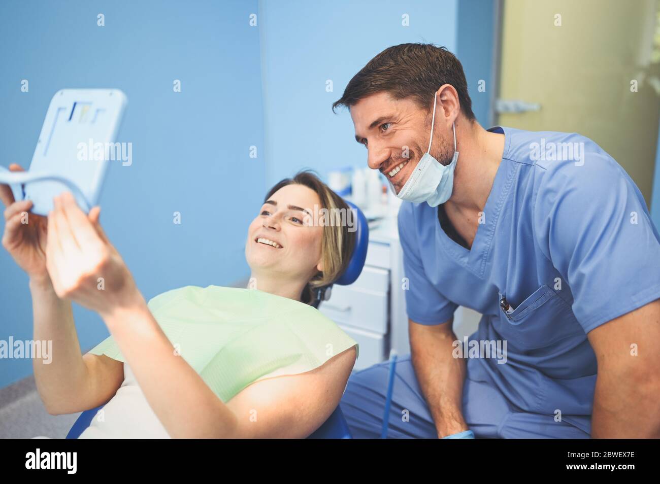Dentist shows the patient the results of treatment with a mirror, examinating teeth with dental ...