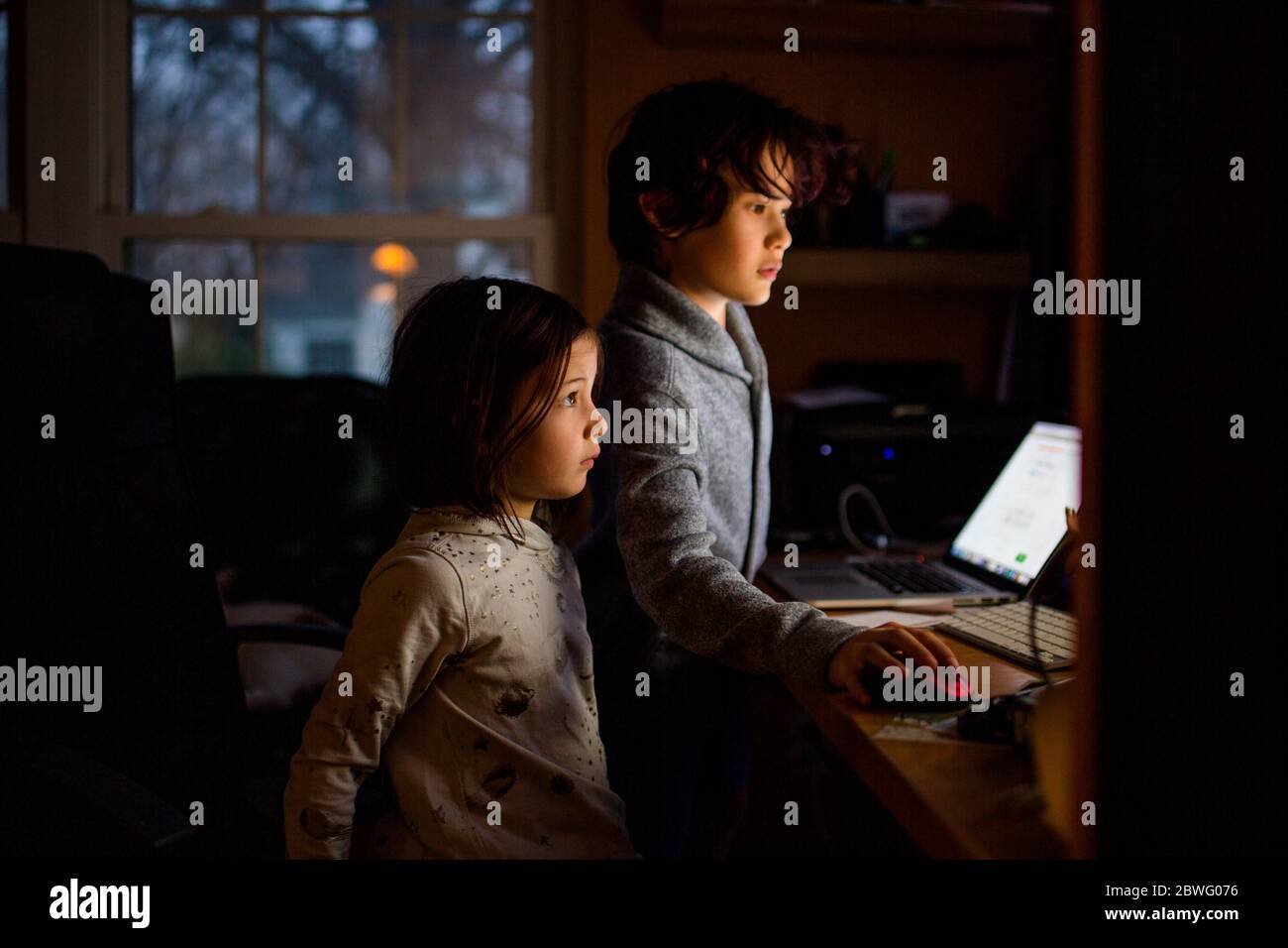 Two children stand in a dark room faces lit up by a computer screen ...