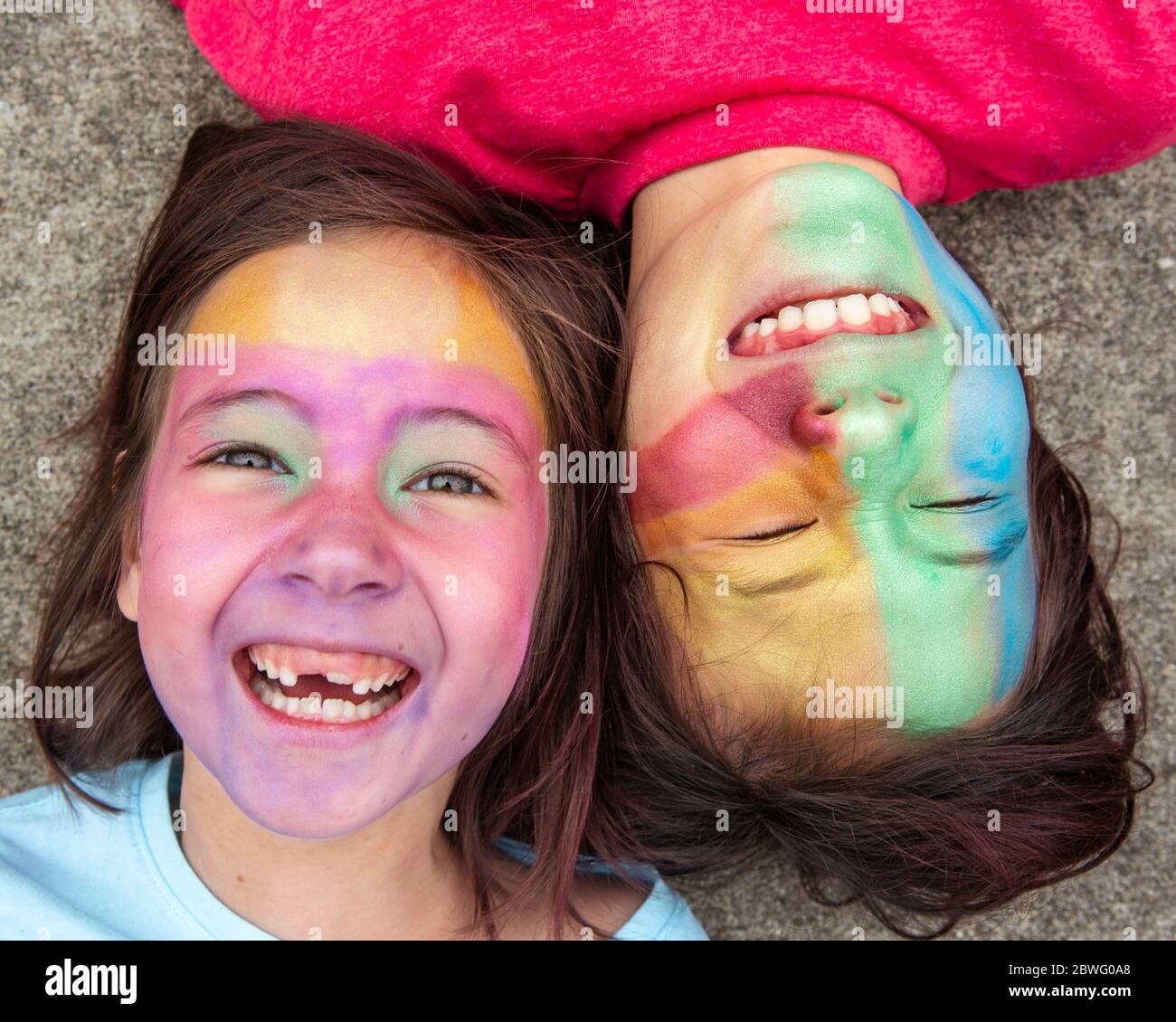 Two children lay down cheek-to-cheek with colorful face paint laughing ...