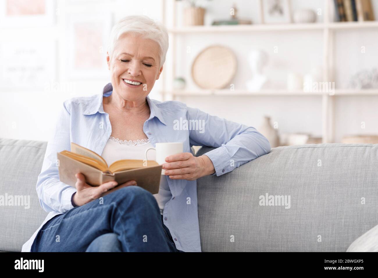 Retirement time. Elderly lady sitting on couch with book and drinking ...