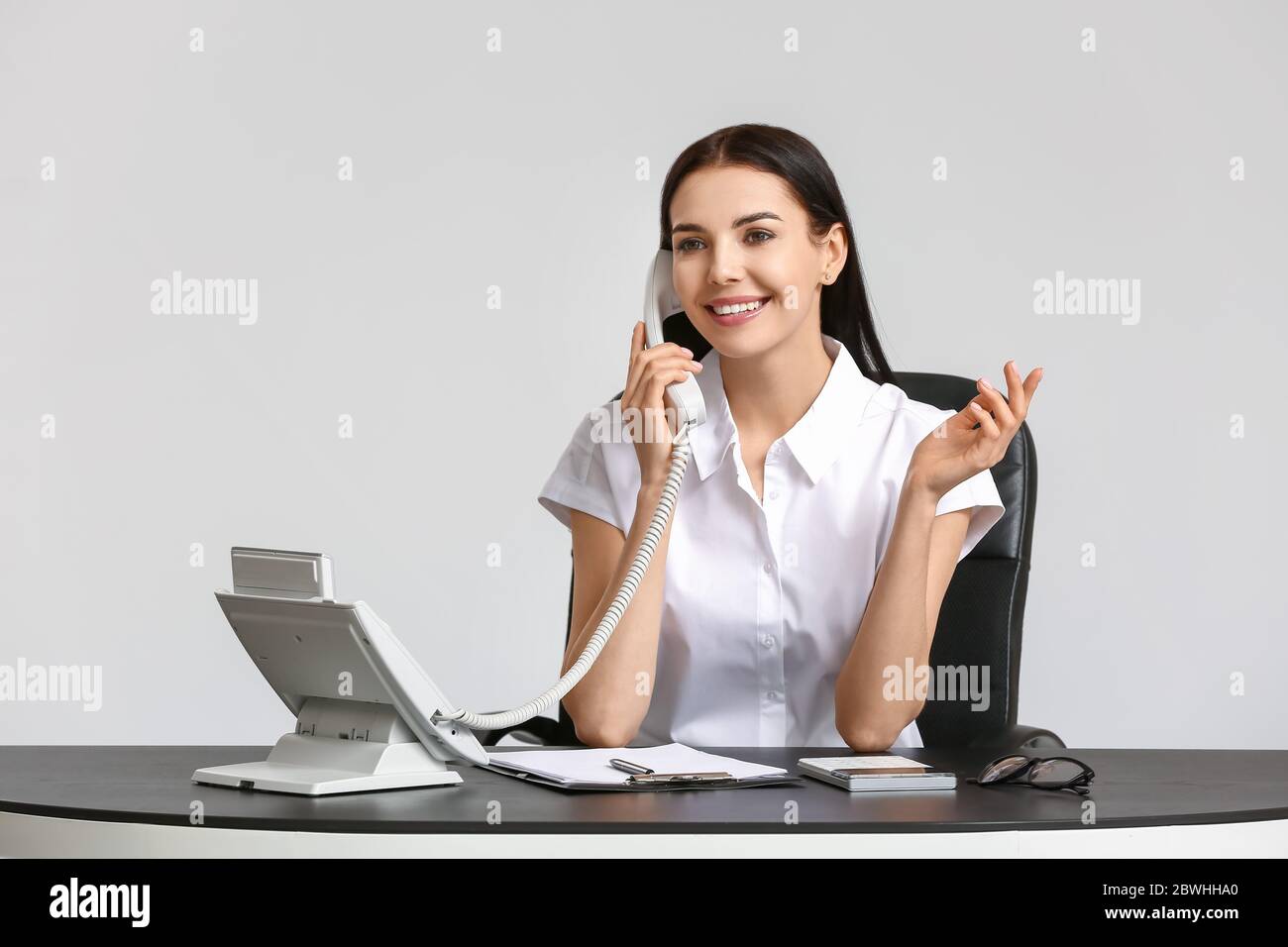 Beautiful young secretary talking by telephone at table on white ...