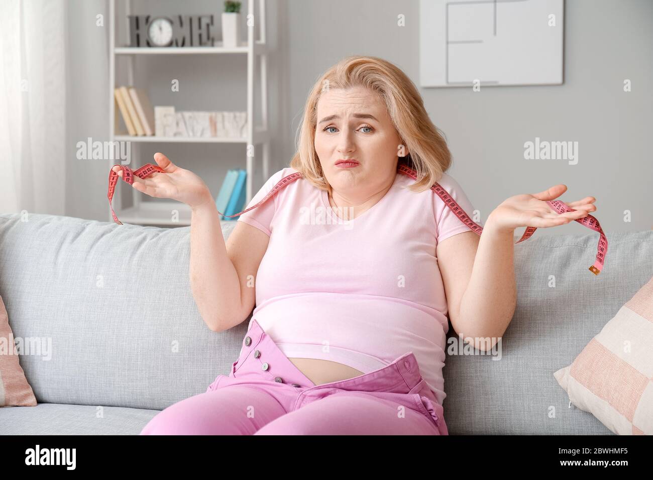 Sad overweight woman in tight clothes and with measuring tape at home ...