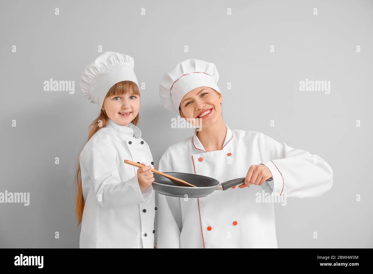 Portrait of female chef and her little daughter on light background ...