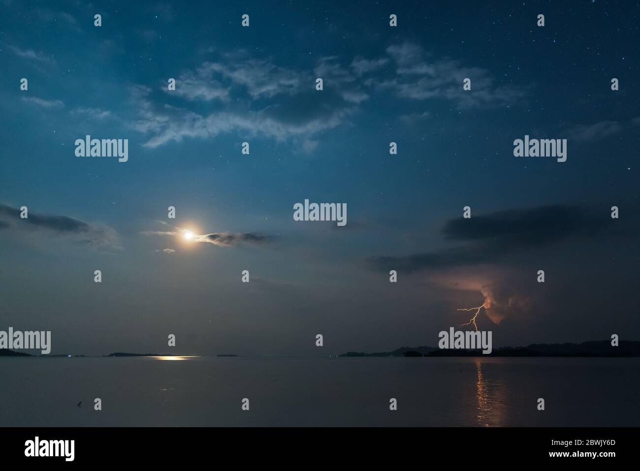 Lightning in the night starry sky with moon reflection over water ...