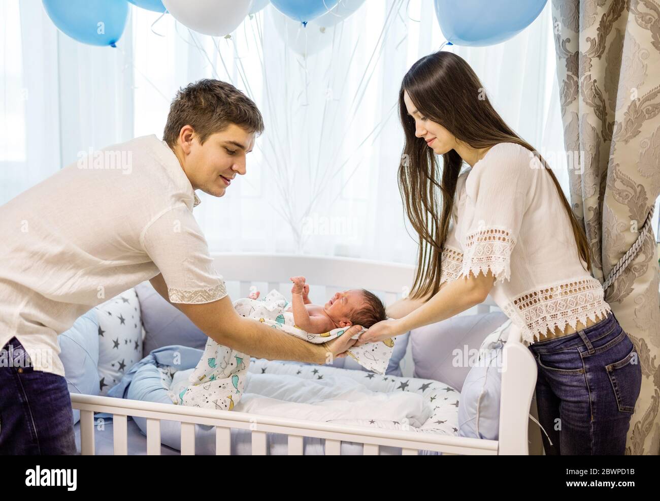 Young couple putting crying newborn son to sleep in white crib next to ...
