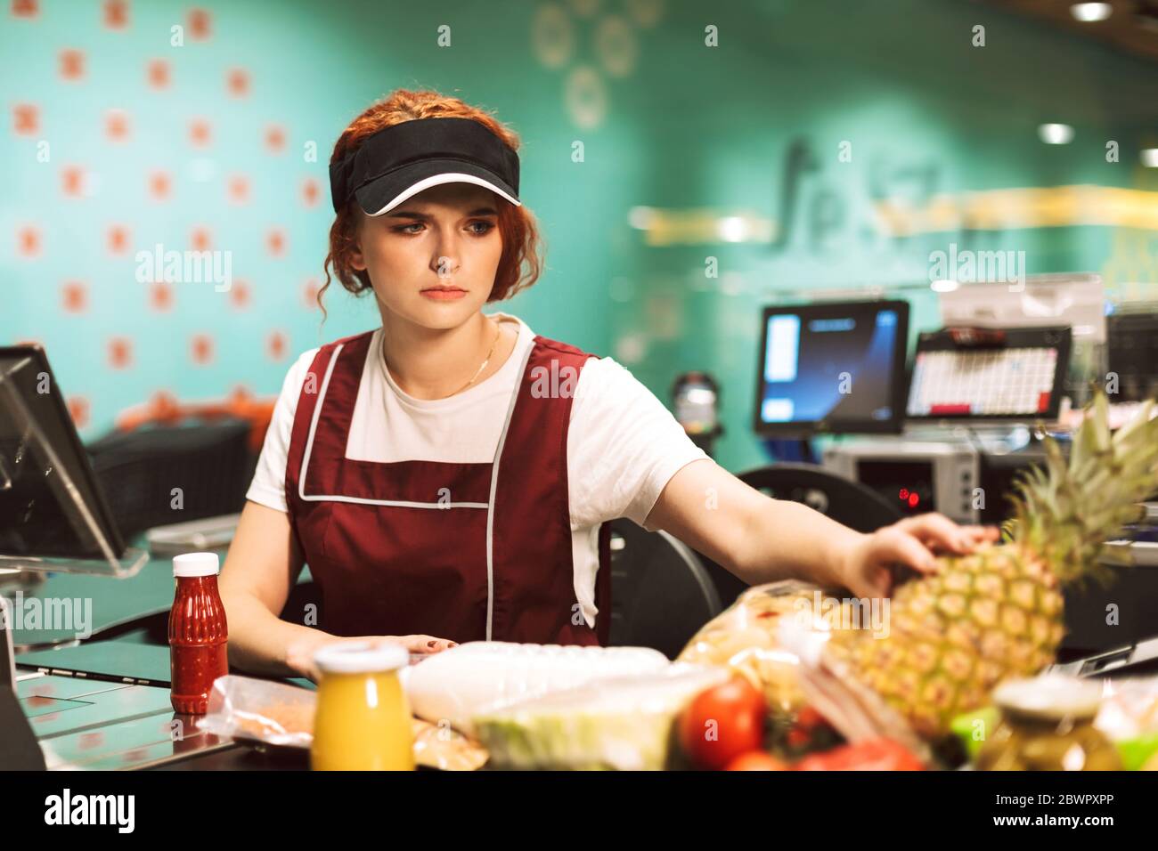 Young upset female cashier in uniform thoughtfully looking on products ...