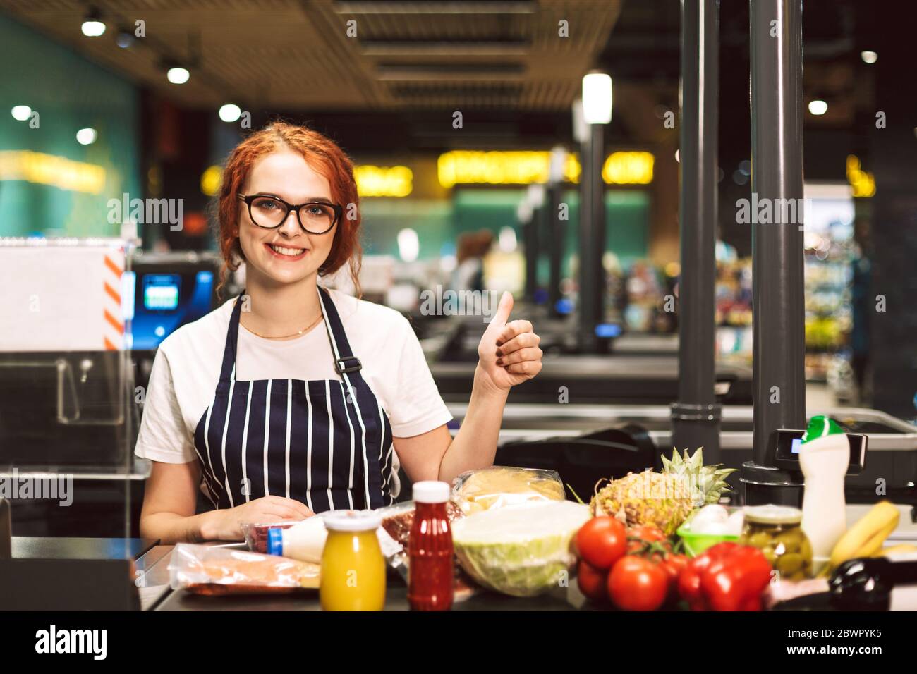Young smiling female cashier in eyeglasses and striped apron with ...
