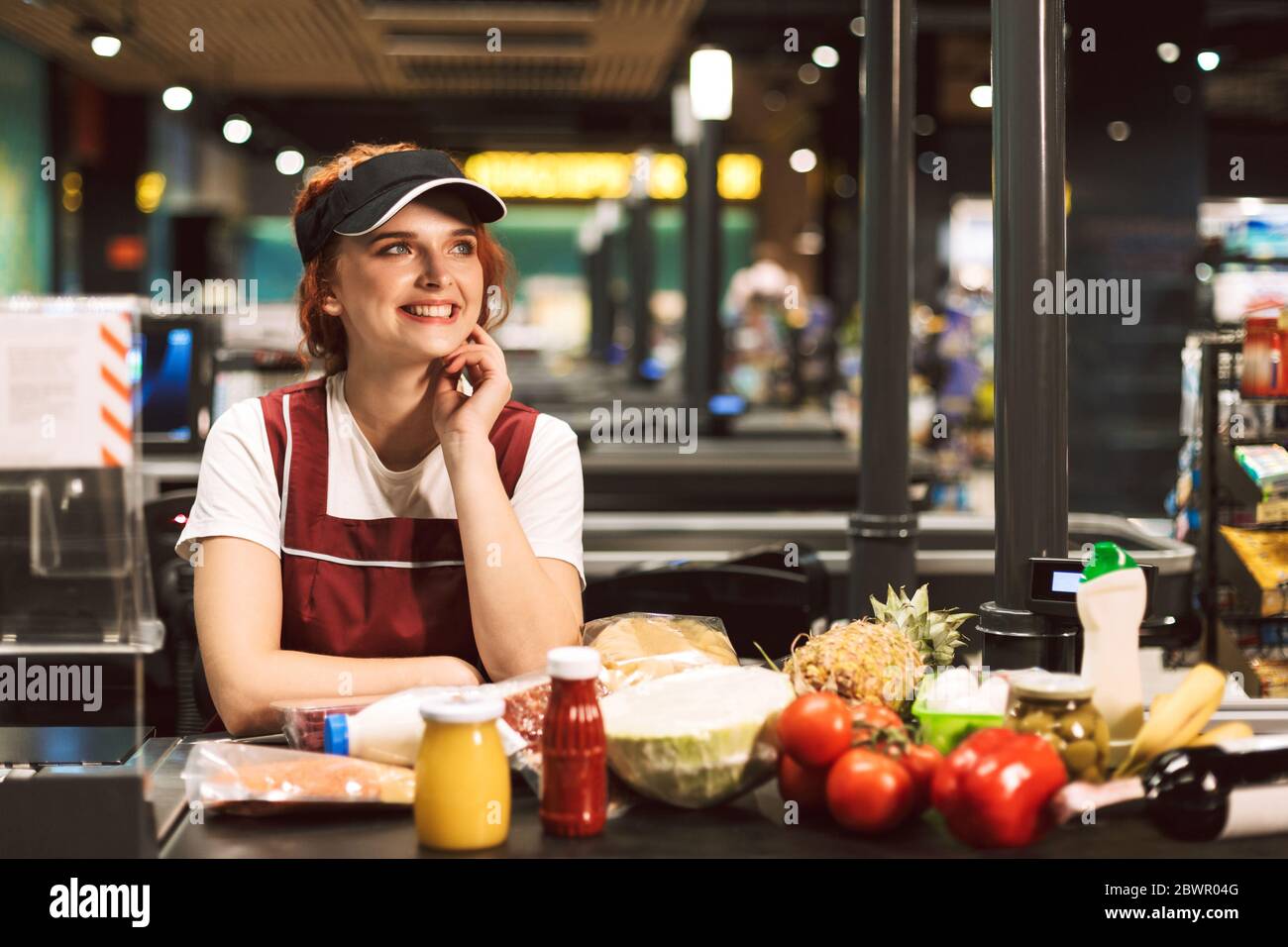 Young cheerful female cashier in uniform joyfully looking aside with ...