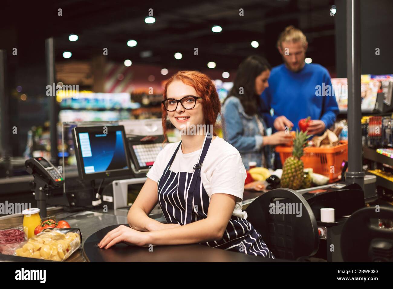 Young smiling cashier in eyeglasses and striped apron happily looking ...