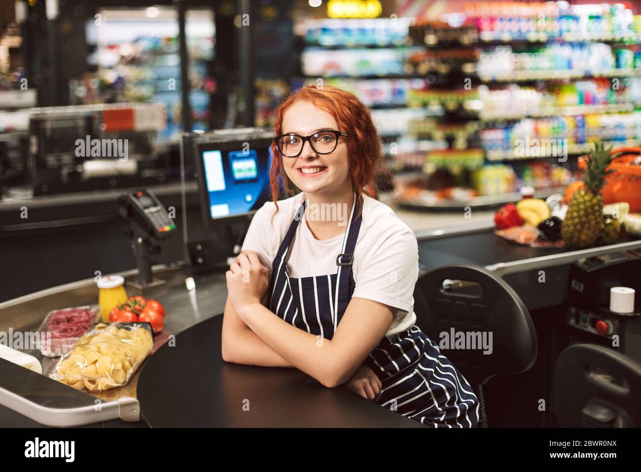 Pretty smiling cashier in eyeglasses and striped apron happily looking ...
