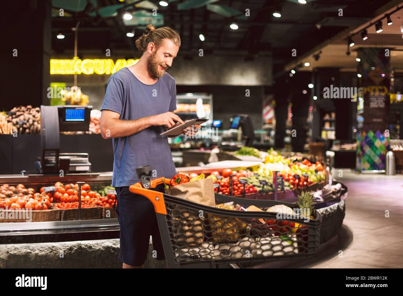 Young smiling man with trolley full of products happily using the ...