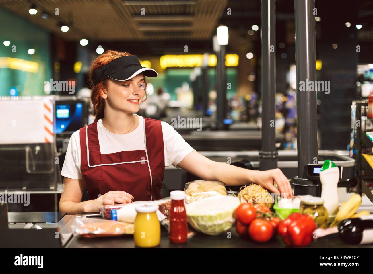 Young female cashier in uniform happily looking aside with products on ...