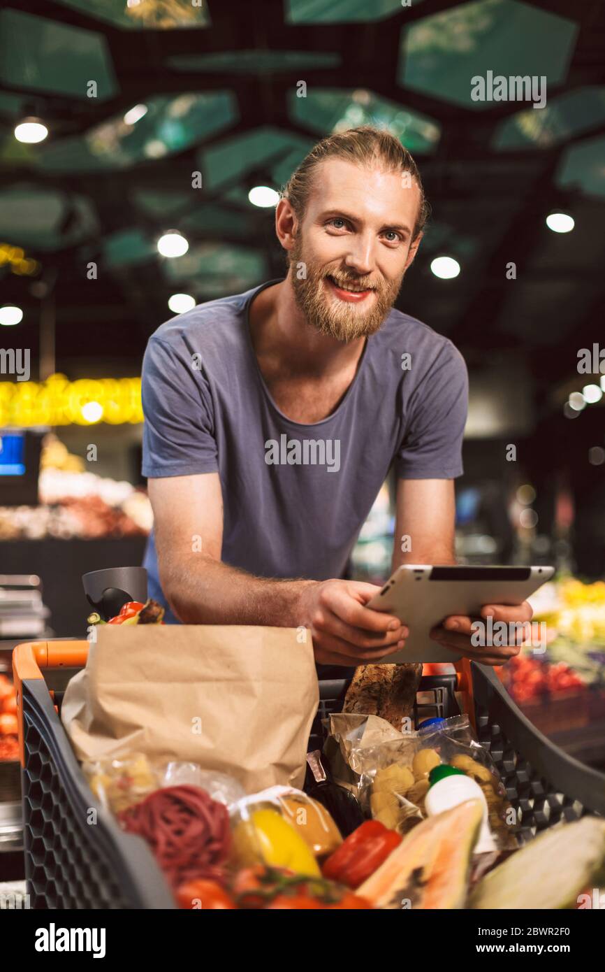 Young guy leaning on trolley full of food joyfully looking in camera ...