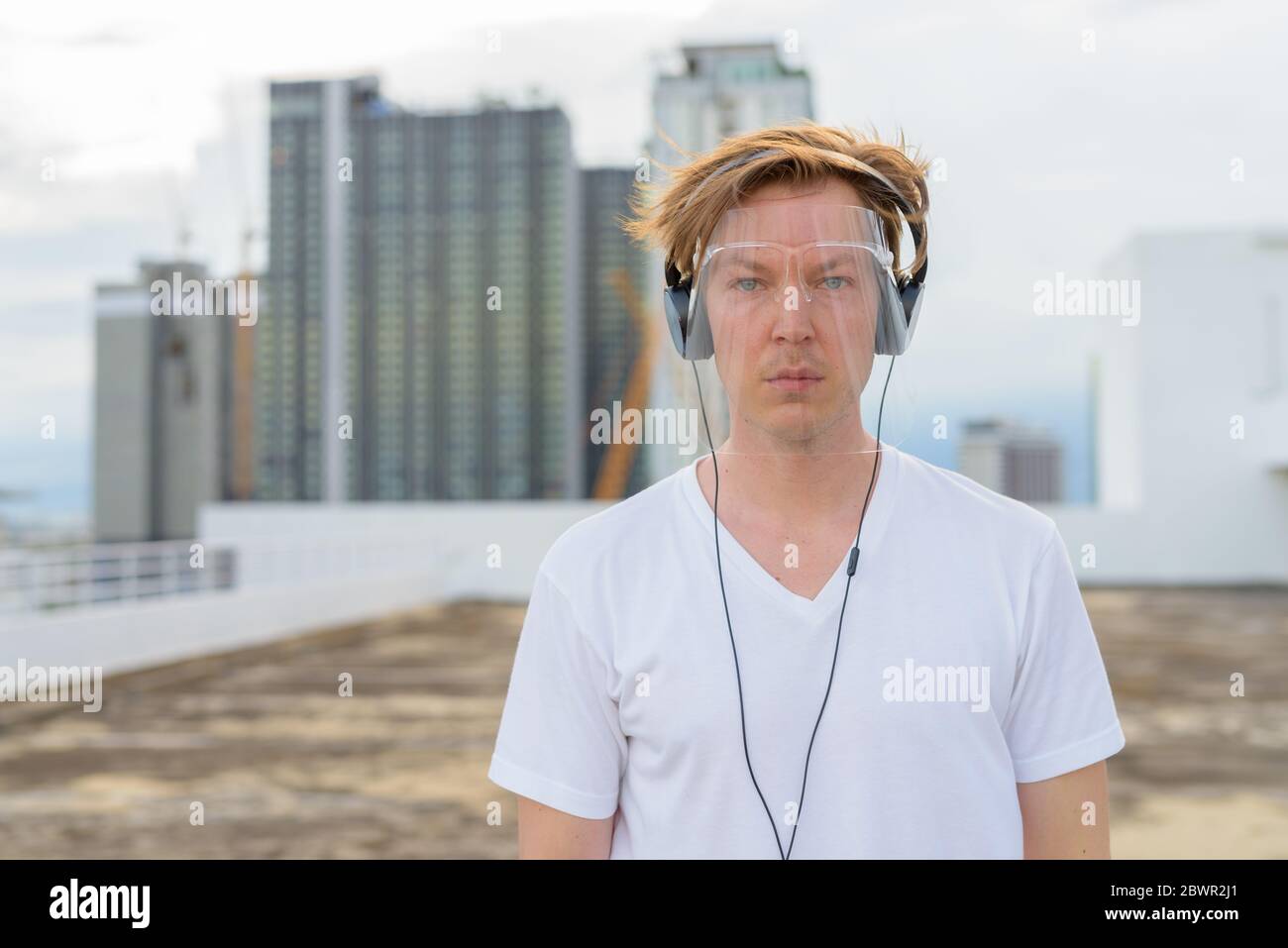 Young handsome man with face shield listening to music at rooftop of ...