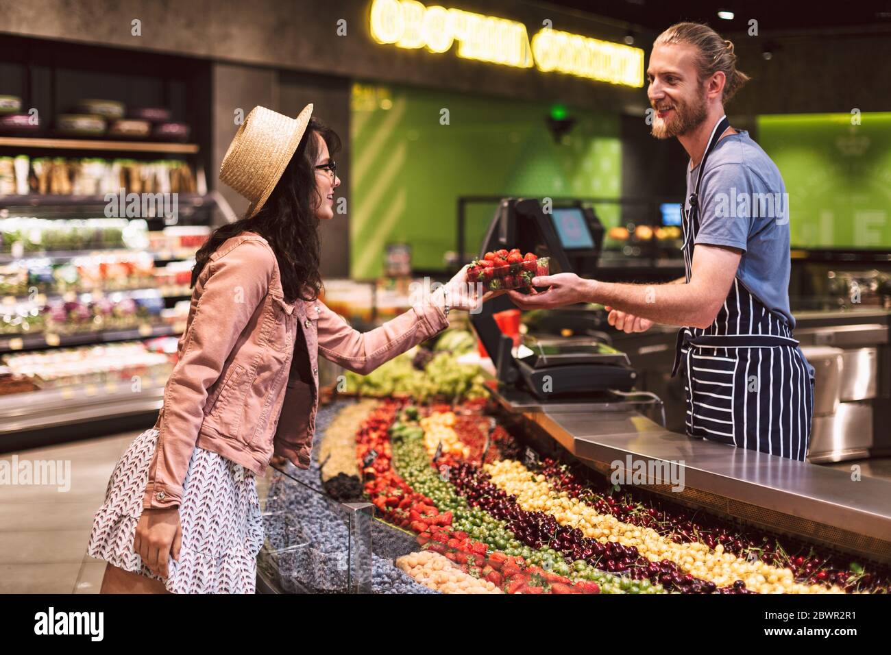 Young smiling seller in apron behind counter happily giving ... Young smiling seller in apron behind counter happily giving ...