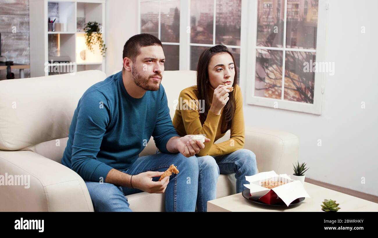Concentrated young couple eating fried chicken while watching tv ...