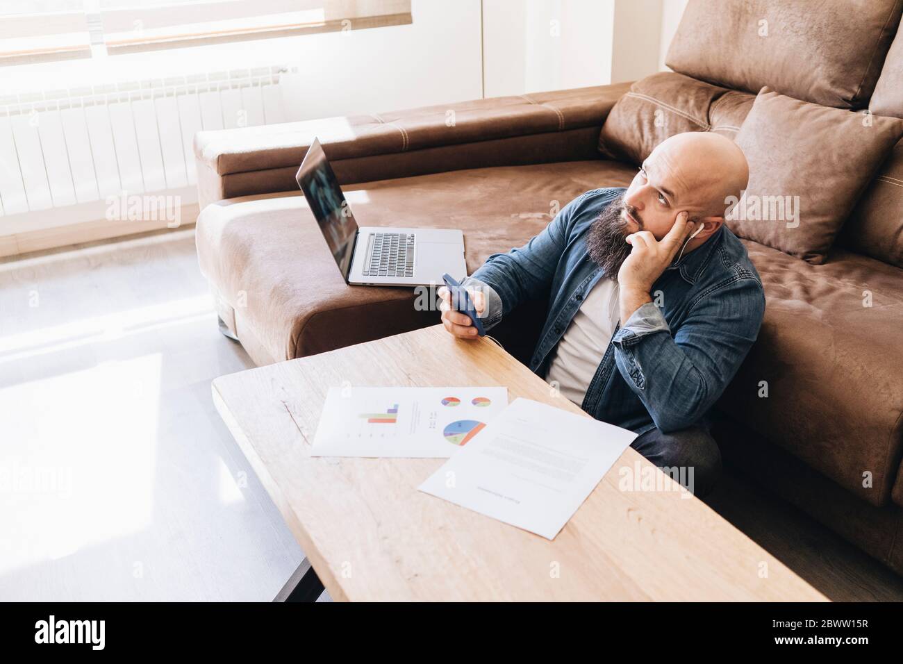 Thoughtful businessman with laptop and documents using smart phone in ...