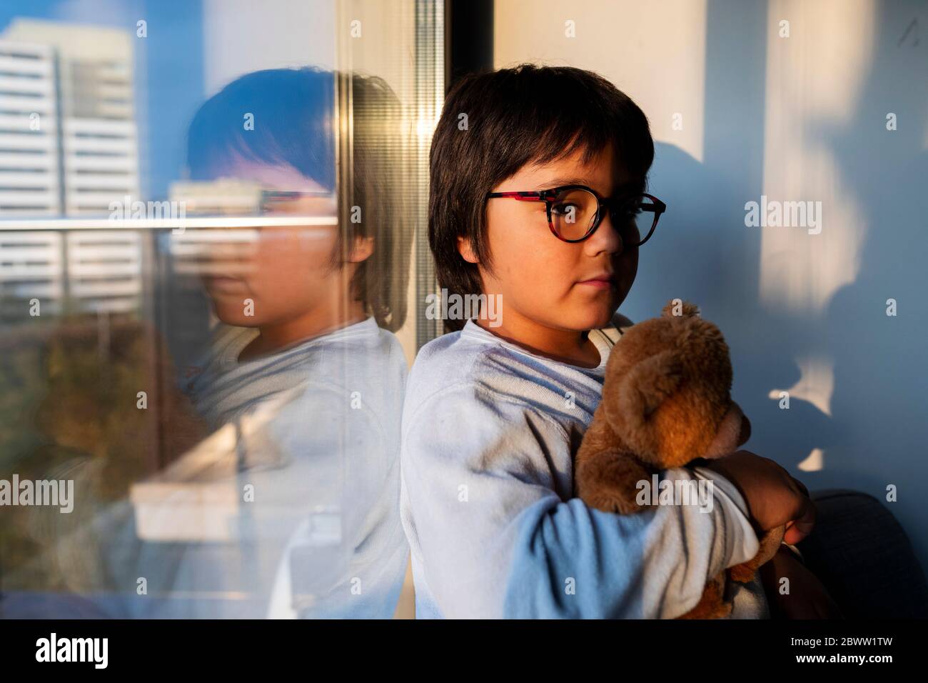 Portrait of serious boy with teddy bear leaning at window in the ...